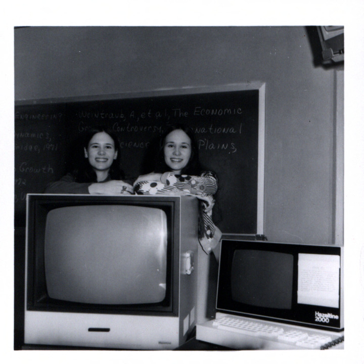 Two female students (?) (twins?) posed over a television and a computer ...